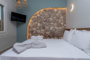 Quarto com parede de pedra, TV de tela plana, cama com roupa de cama branca e manta cinza.