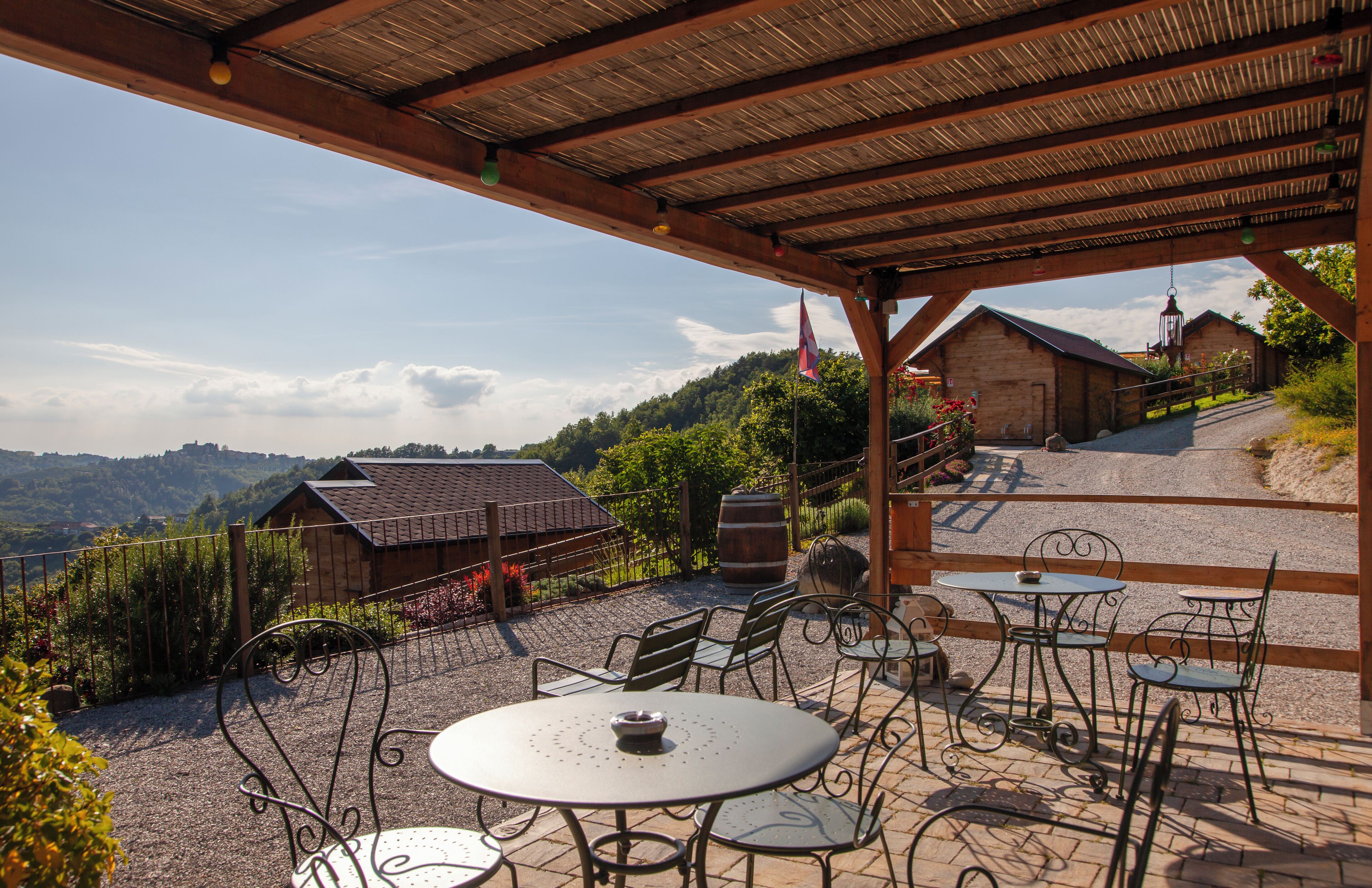 Uma área de estar coberta ao ar livre, com mesas e cadeiras, uma pérgola e vista para uma colina com edifícios.