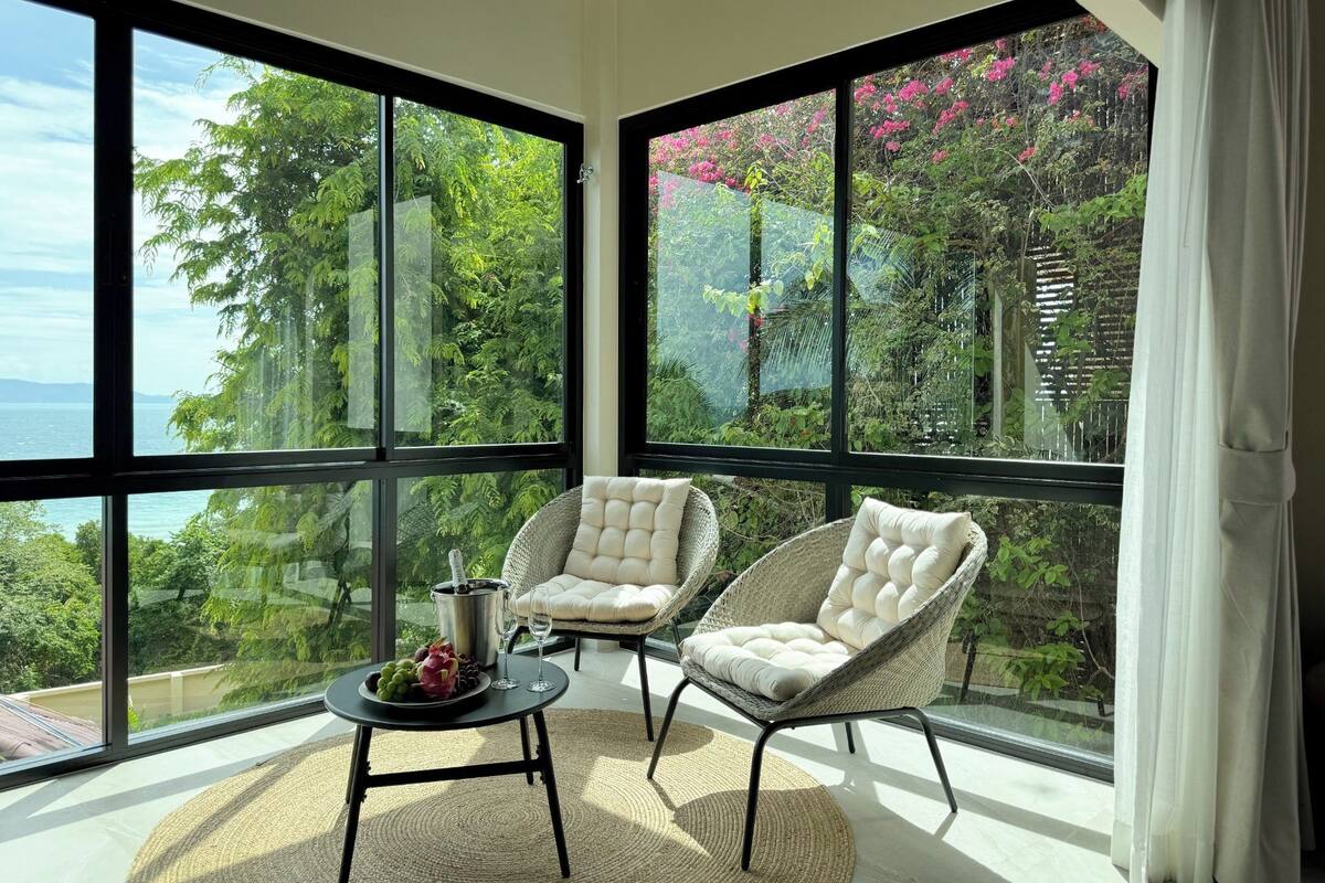 Quarto com janelas amplas, cadeiras de vime, uma mesinha com bebida e frutas, e vista para o verde e a água.