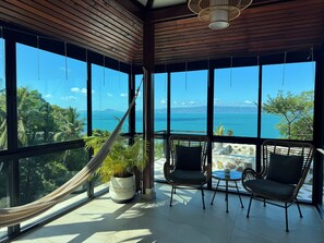 Quarto com grandes janelas, uma rede, duas cadeiras de vime, uma mesa e um vaso com planta.