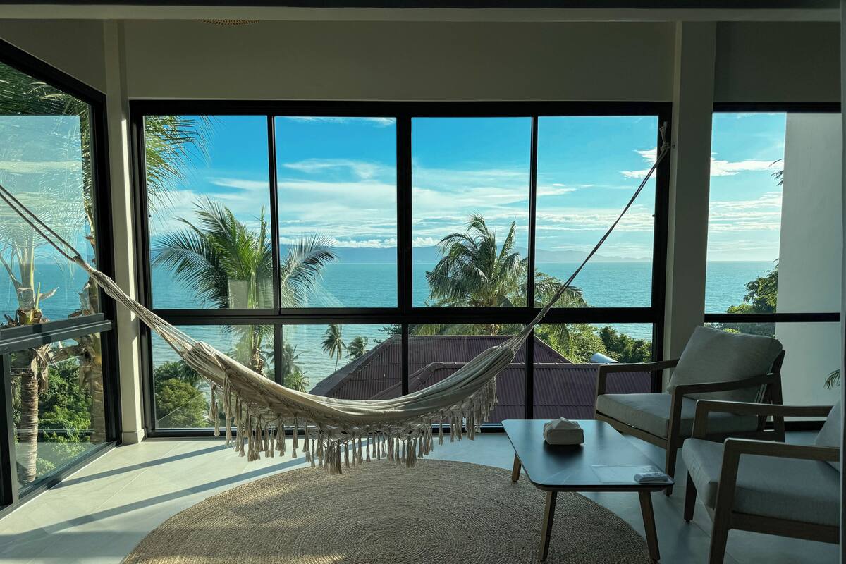 Quarto com janelas amplas, uma rede, um tapete redondo e vista para o oceano.