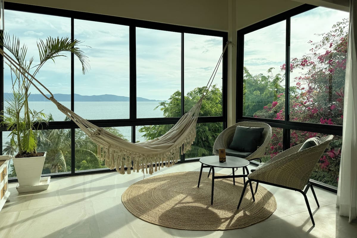 Quarto amplo com grandes janelas que oferecem vista para o mar e montanhas. Há uma rede, duas cadeiras de vime e um tapete redondo tecido.
