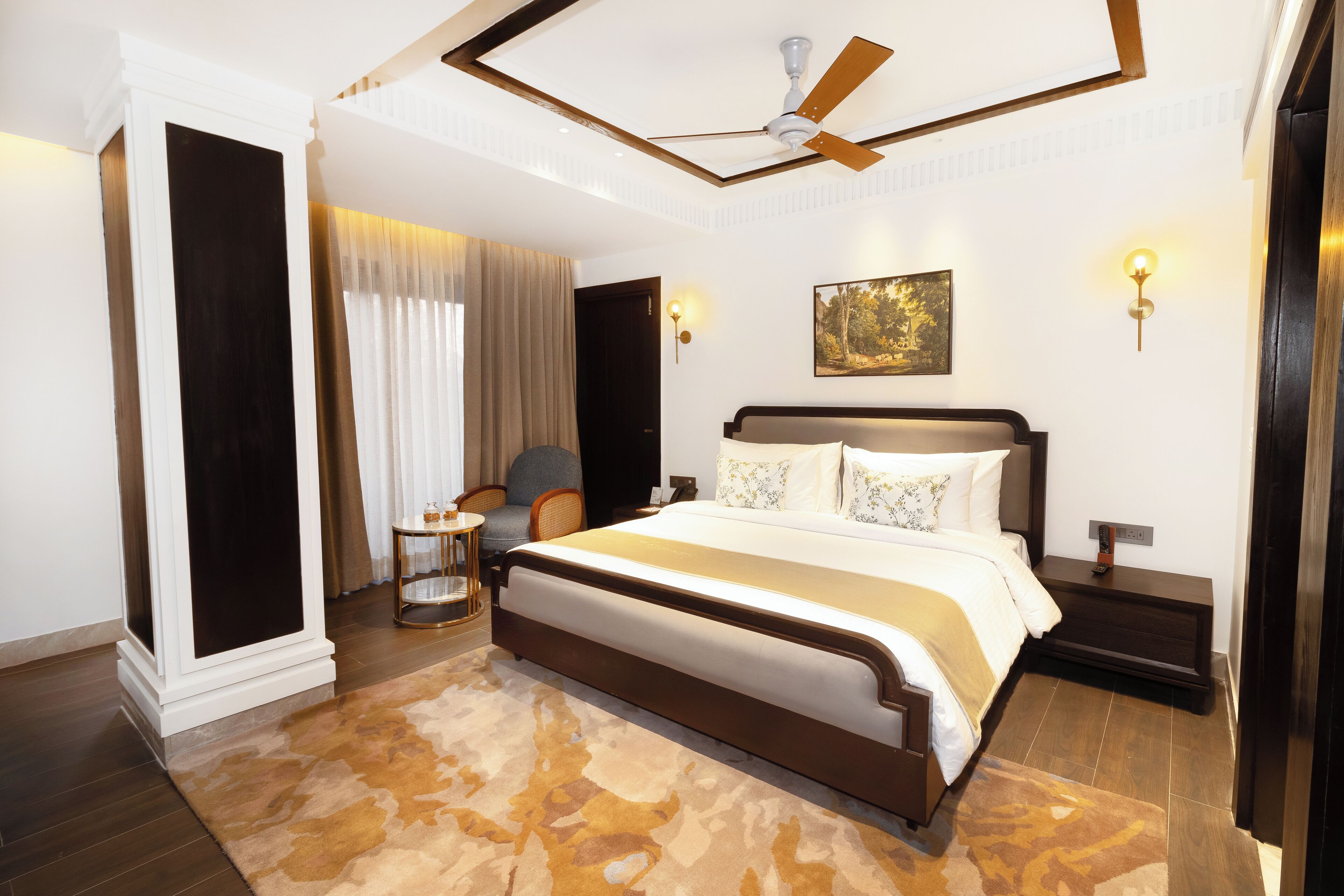 Quarto de hotel com uma cama grande, um ventilador de teto, mesa de cabeceira, cadeira e janela com cortinas.