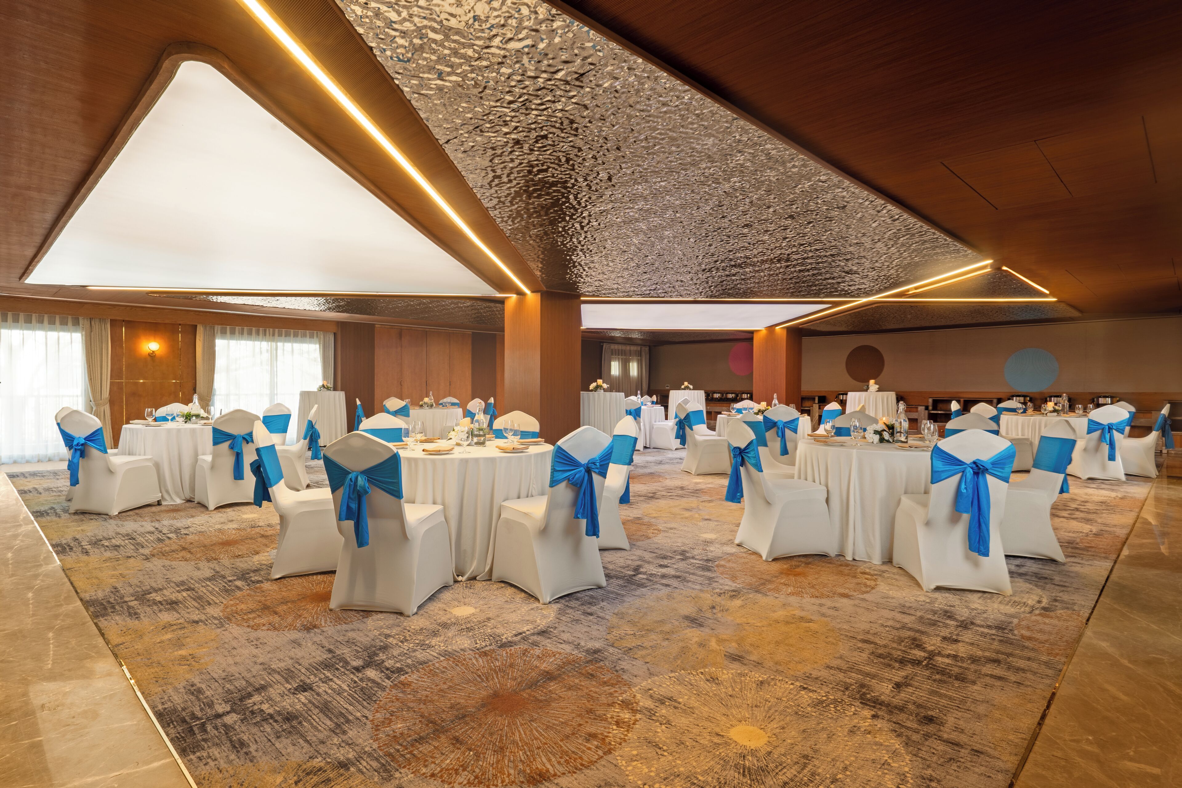 Uma sala de banquetes com mesas redondas, cadeiras brancas com fitas azuis e um tapete estampado.
