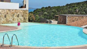 Piscina com paredes de pedra, vista clara do mar e paisagem montanhosa ao fundo.