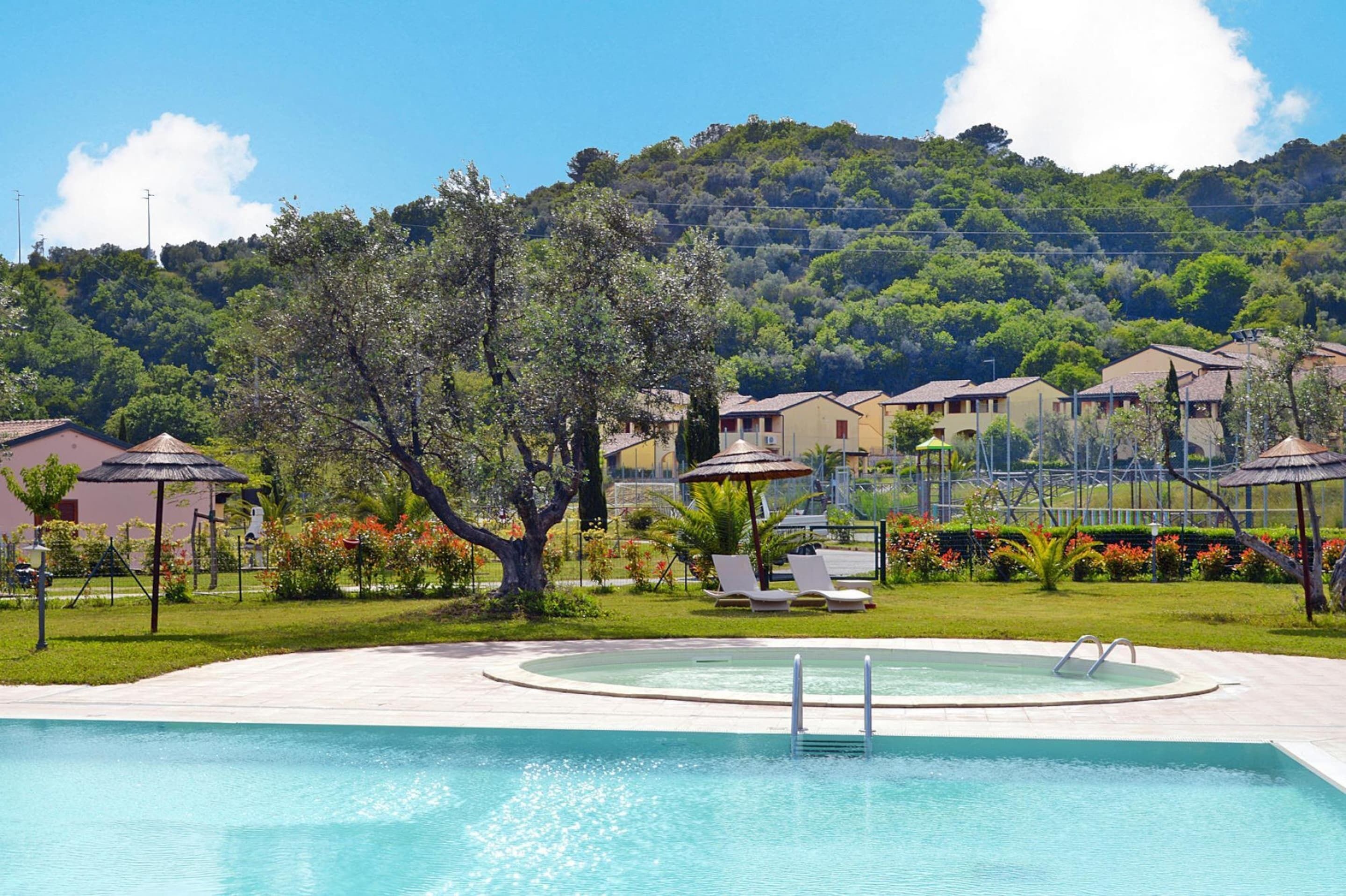 Piscina com espreguiçadeiras e guarda-sóis, cercada por vegetação e edifícios ao fundo.