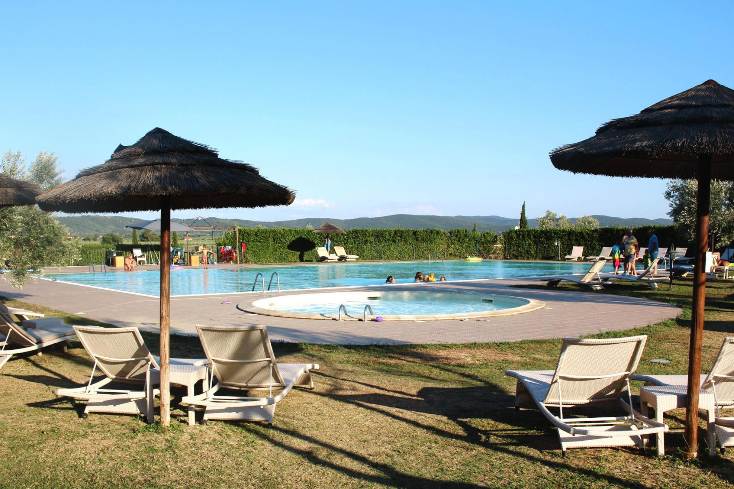 Piscina com espreguiçadeiras e guarda-sóis de palha, rodeada por vegetação e colinas ao fundo.