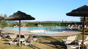 Piscina com espreguiçadeiras e guarda-sóis de palha, rodeada por vegetação e colinas ao fundo.