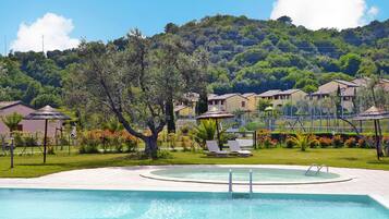 Uma piscina com espreguiçadeiras e guarda-sóis, cercada por vegetação e casas ao fundo.