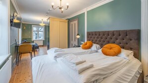 Quarto de hotel com duas camas, área de estar com uma cadeira e uma mesa, uma janela grande e uma porta que dá para outro cômodo.