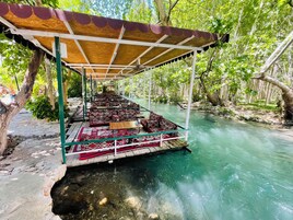 Uma área de estar coberta com bancos e mesas, à beira de um rio calmo, rodeada por árvores.