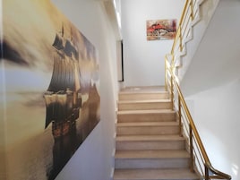 Uma escadaria com degraus bege e corrimãos dourados, que leva a um patamar com um quadro emoldurado.