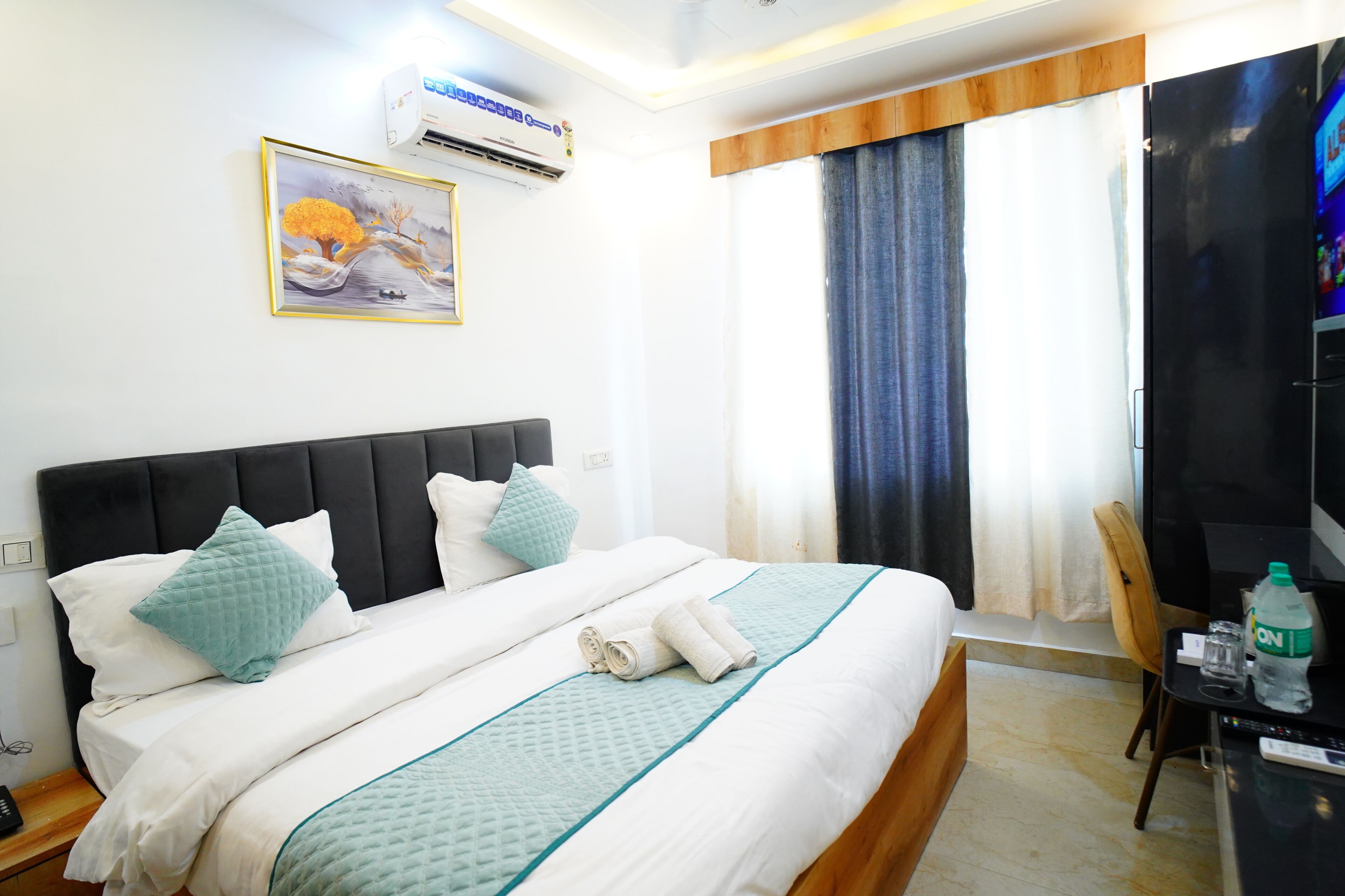 Quarto de hotel com cama, televisão, escrivaninha e janela com cortinas.