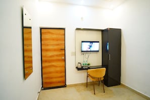 Quarto com porta de madeira, televisão fixada na parede, mesa pequena com cadeira e frigorífico.