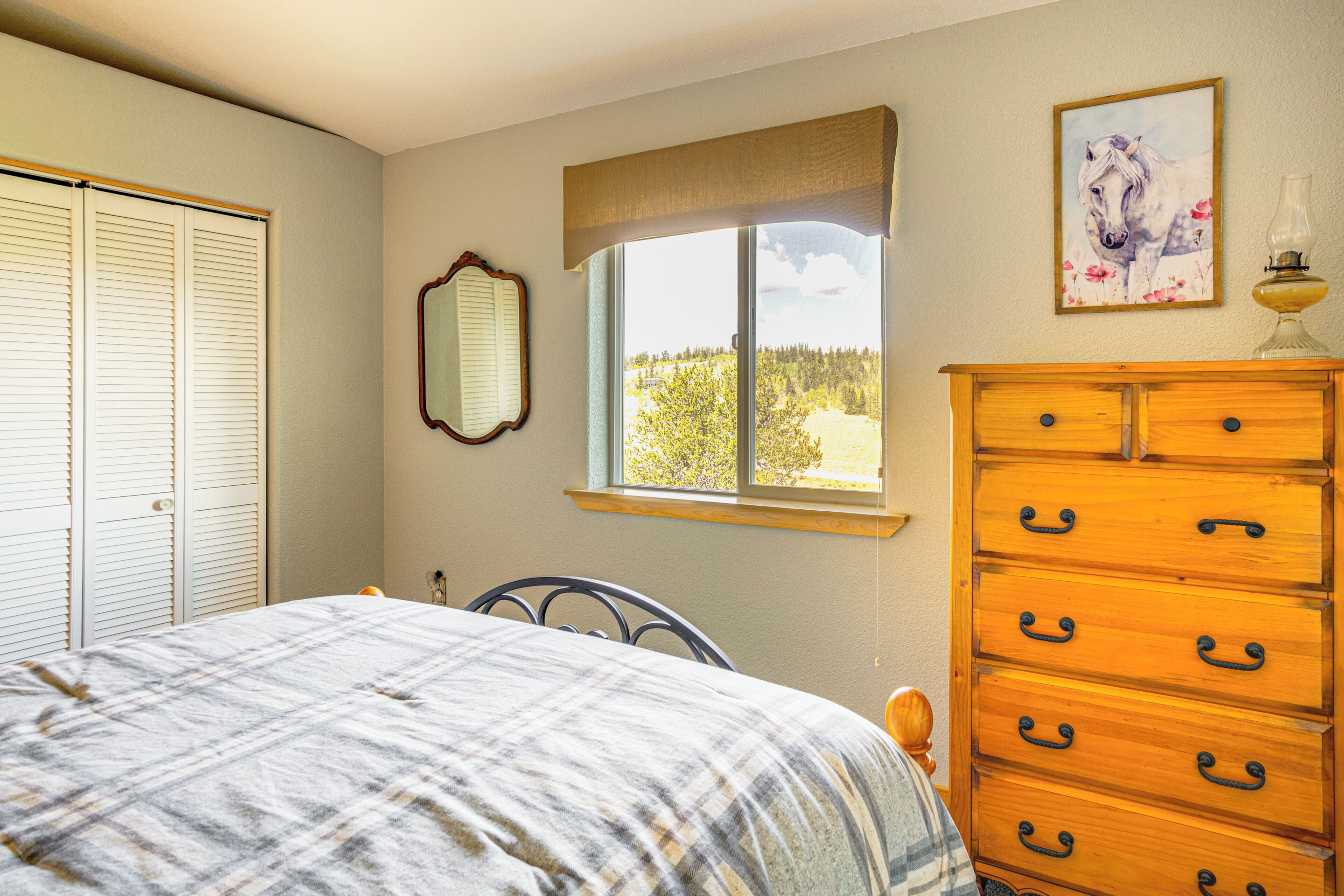 Um quarto com cama, cômoda, janela com vista para árvores e espelho na parede.