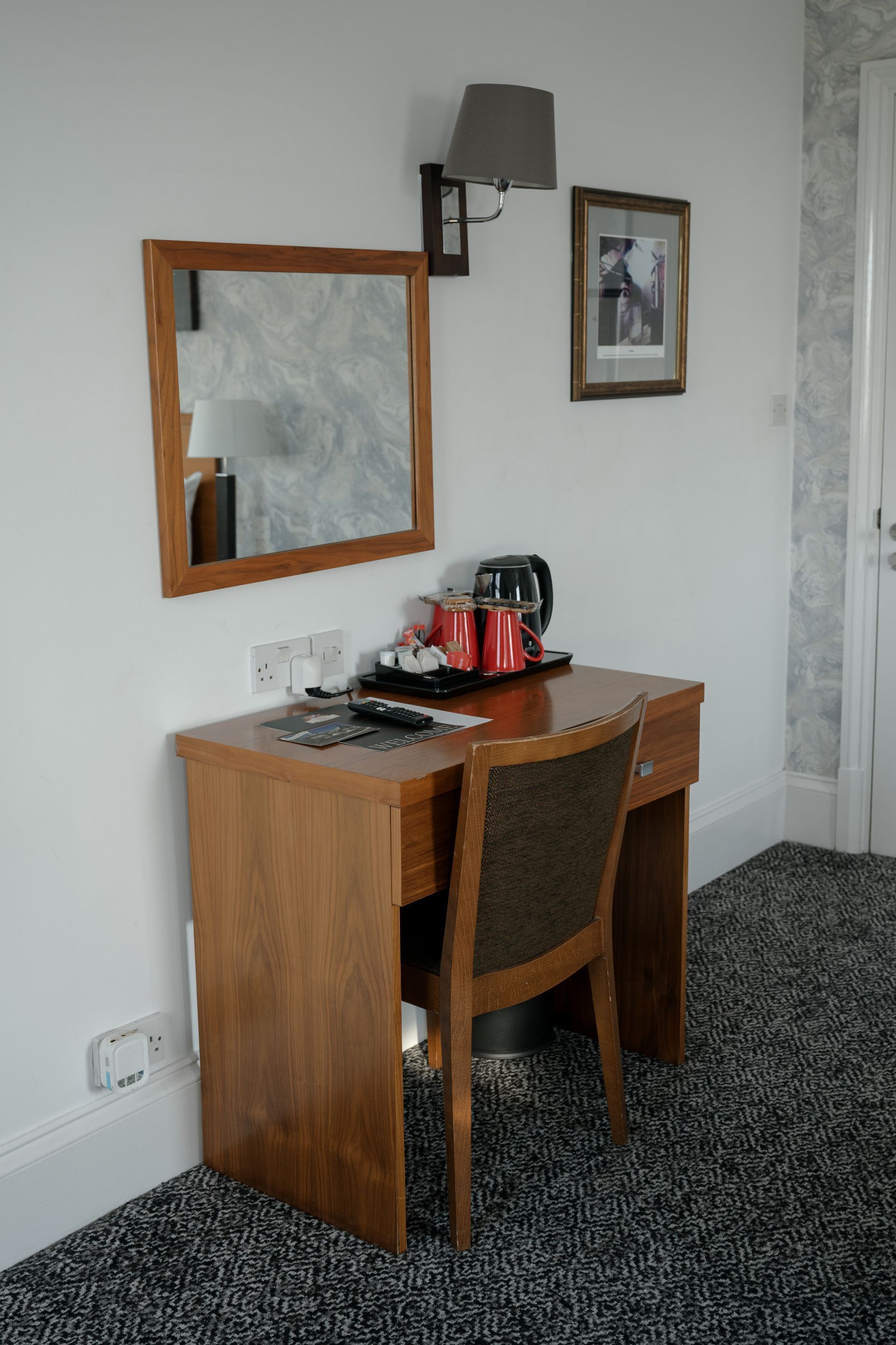 Uma mesa de madeira com uma cadeira, uma lâmpada, um quadro emoldurado e uma cafeteira.