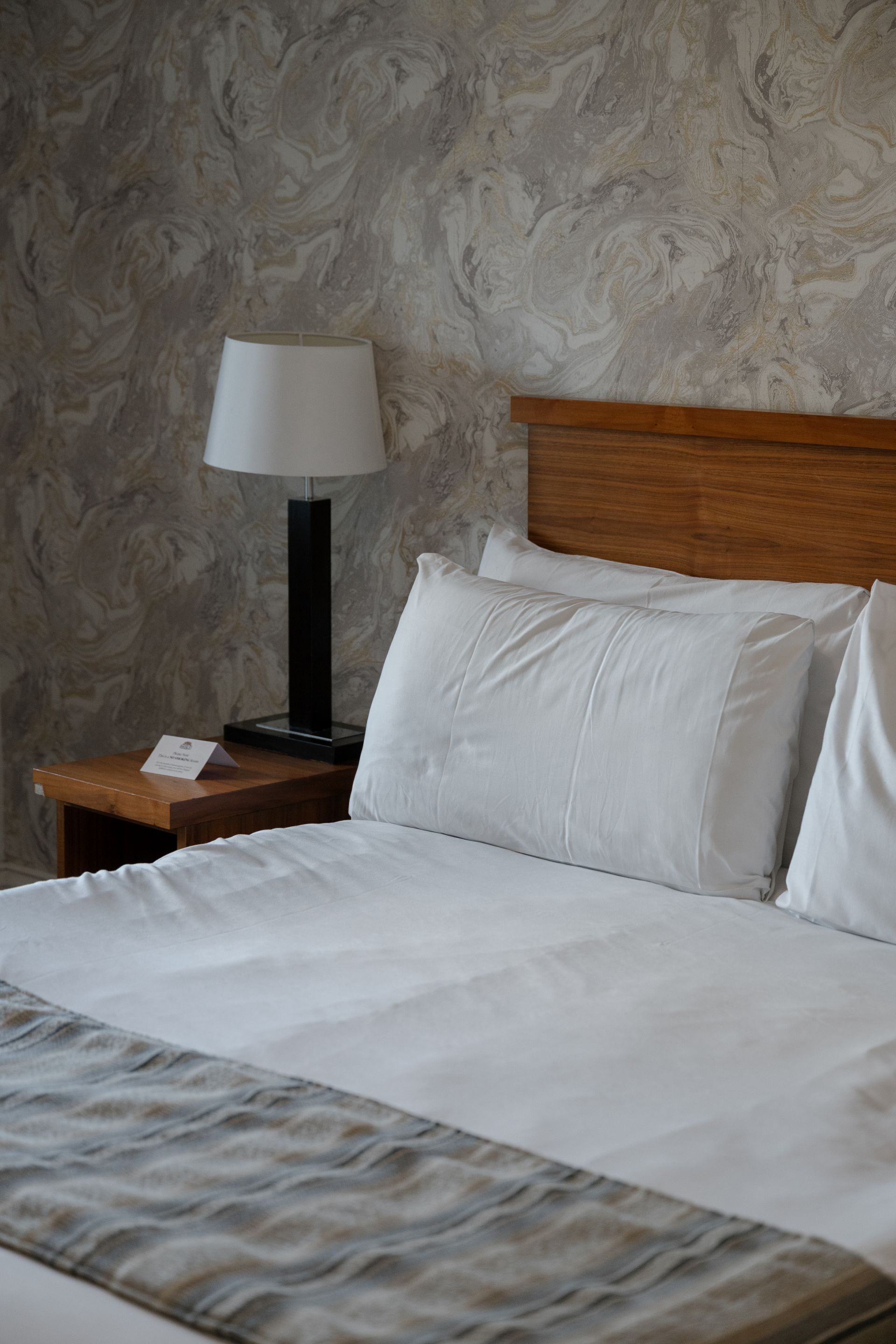 Cama bem arrumada com lençóis brancos e cabeceira de madeira, mesa de cabeceira com abajur e parede com padrão marmorizado.