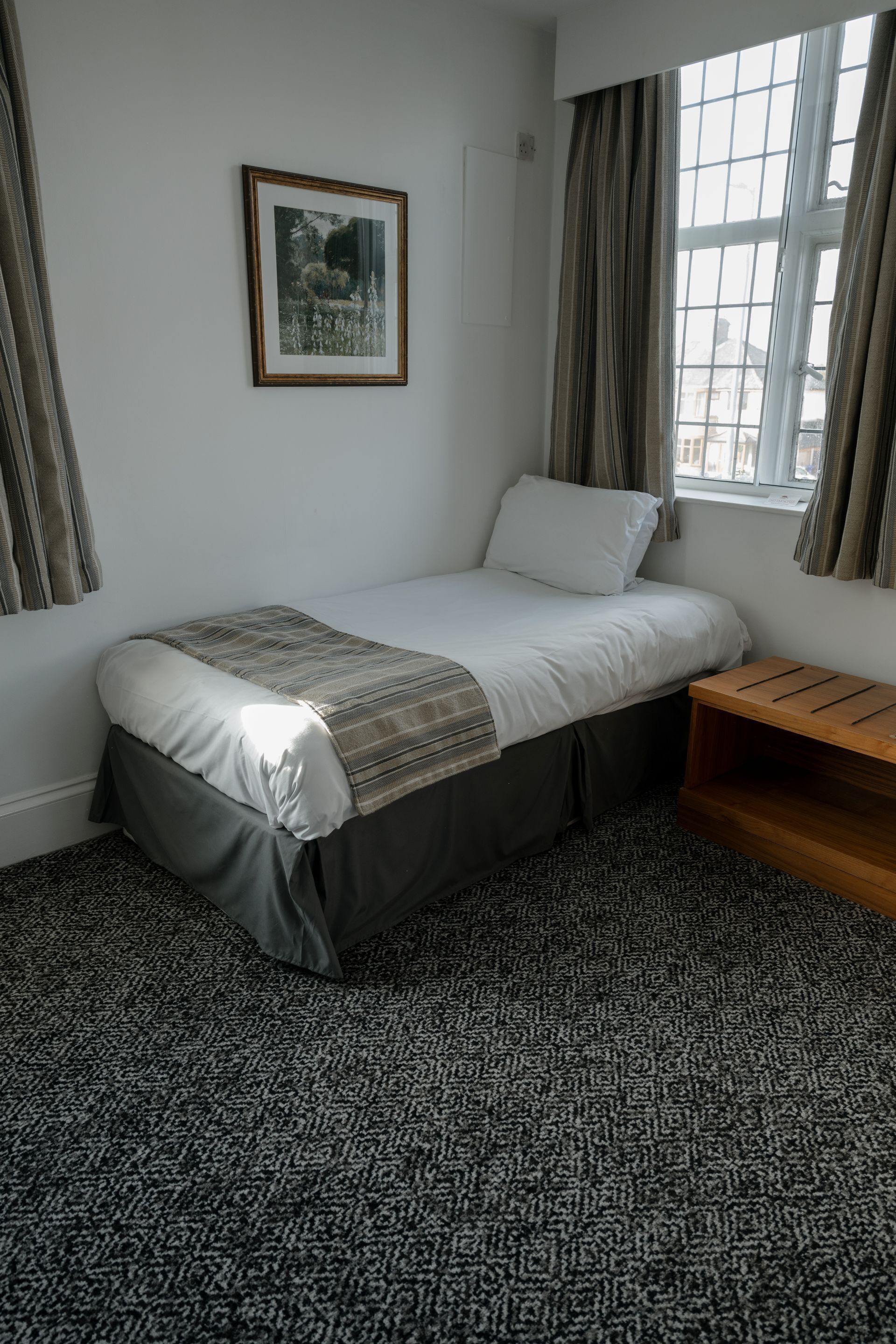 Uma cama de solteiro com um cobertor listrado, uma pequena mesa de cabeceira de madeira, um quadro na parede e uma janela com cortinas.