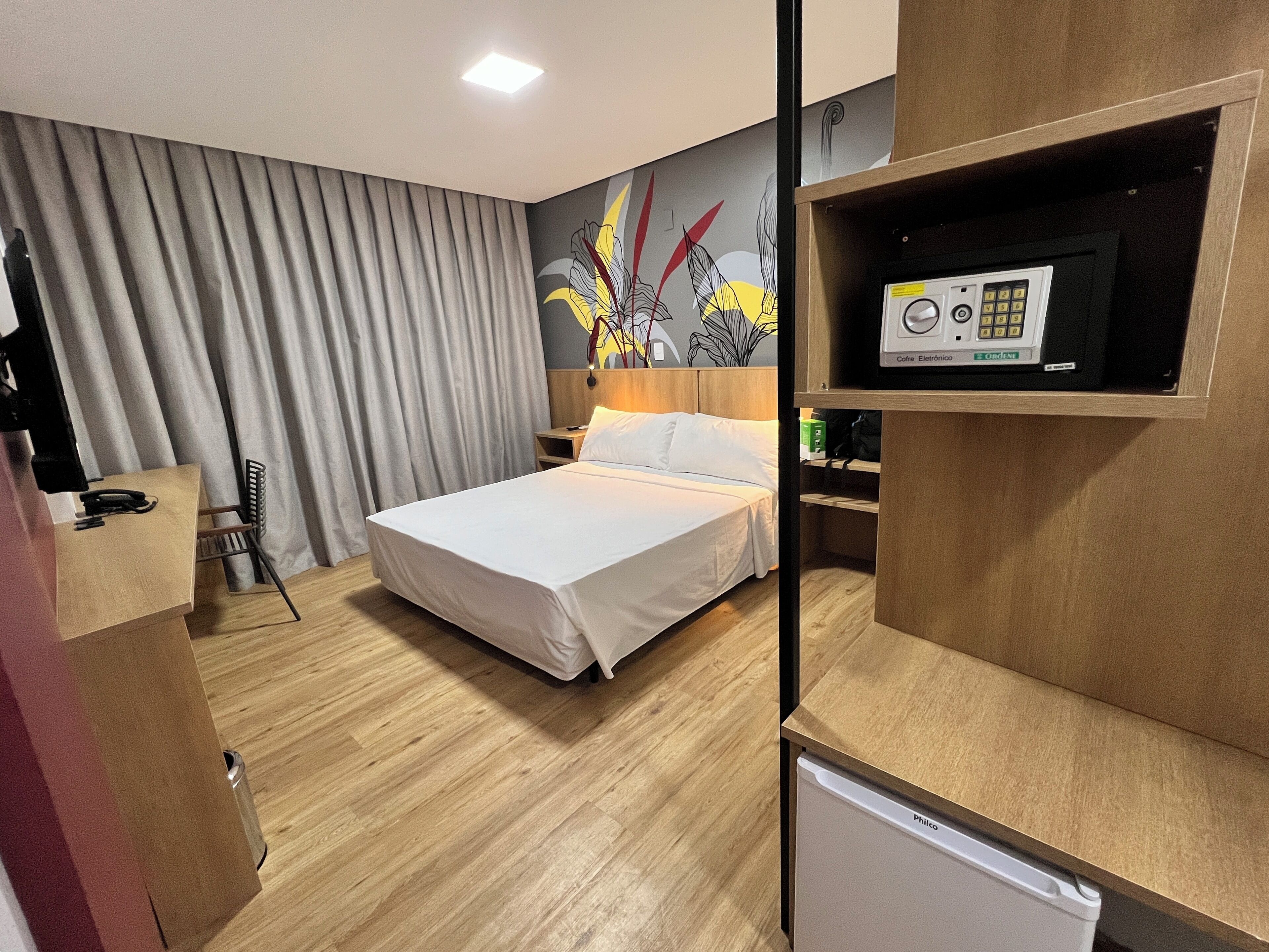 Quarto de hotel com cama, secretária, cadeira, minibar e televisão na parede.