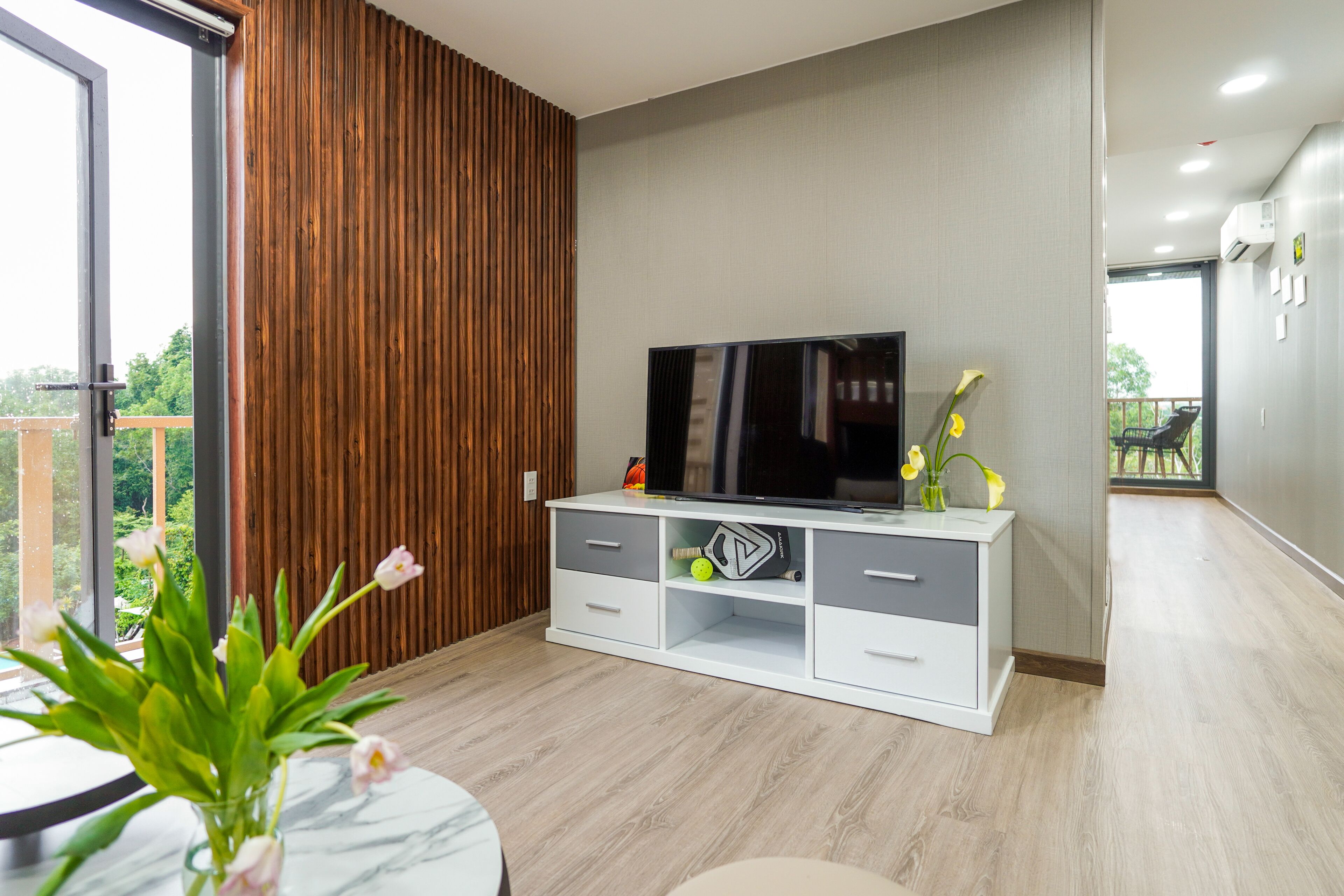 Sala de estar moderna com televisão de tela plana em um armário branco, parede de detalhes em madeira e vista para o exterior.