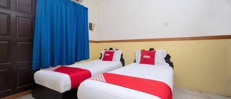 Quarto de hotel com duas camas, cada uma com roupa de cama branca e detalhes vermelhos, uma porta de madeira escura fechada e uma cortina azul.