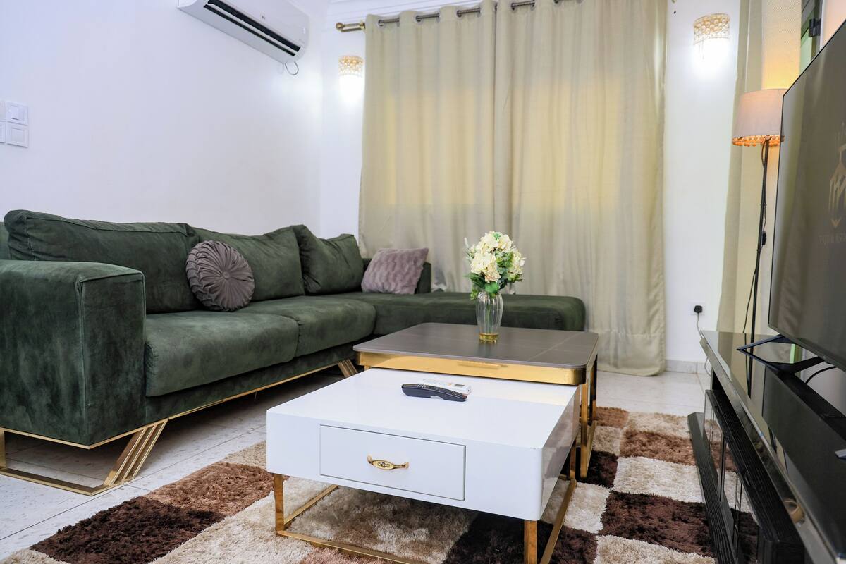 Sala de estar com um sofá verde, uma mesa de centro branca com um controle remoto e uma TV sobre um suporte.