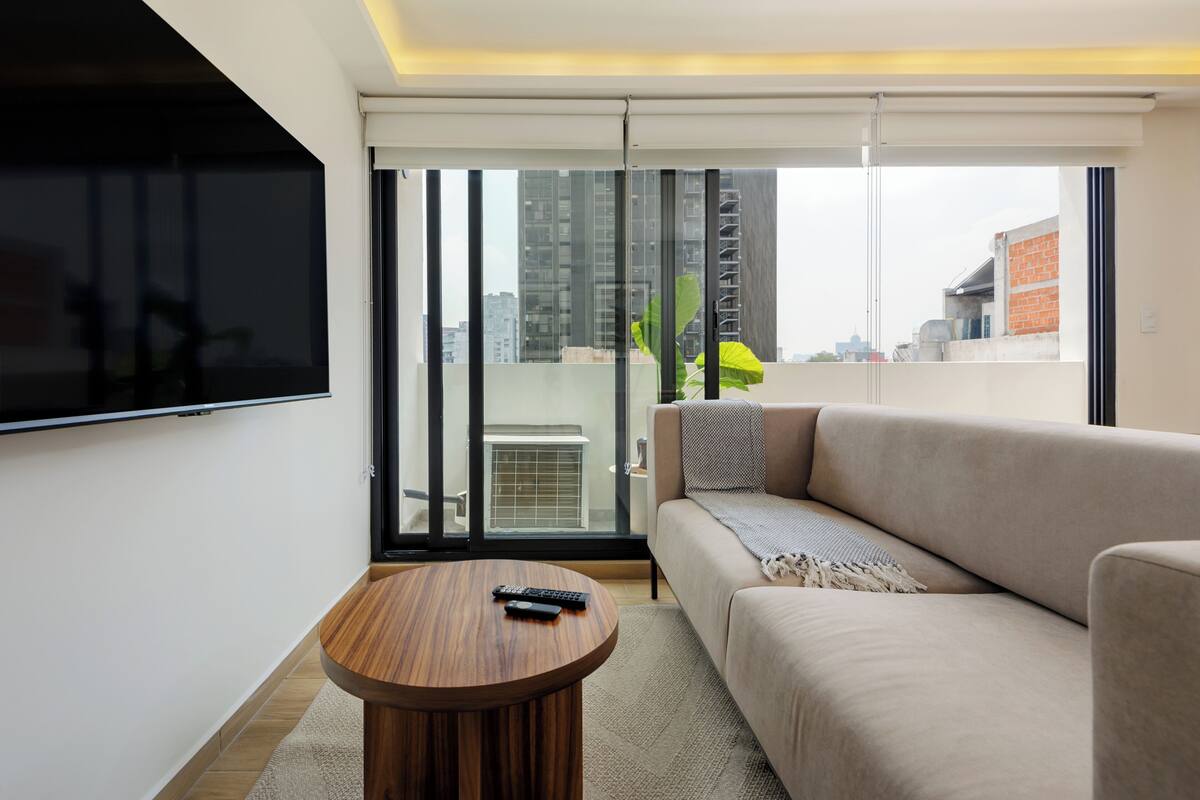 Uma sala de estar moderna com um sofá, uma mesa de centro redonda de madeira e uma televisão de tela plana fixada na parede.