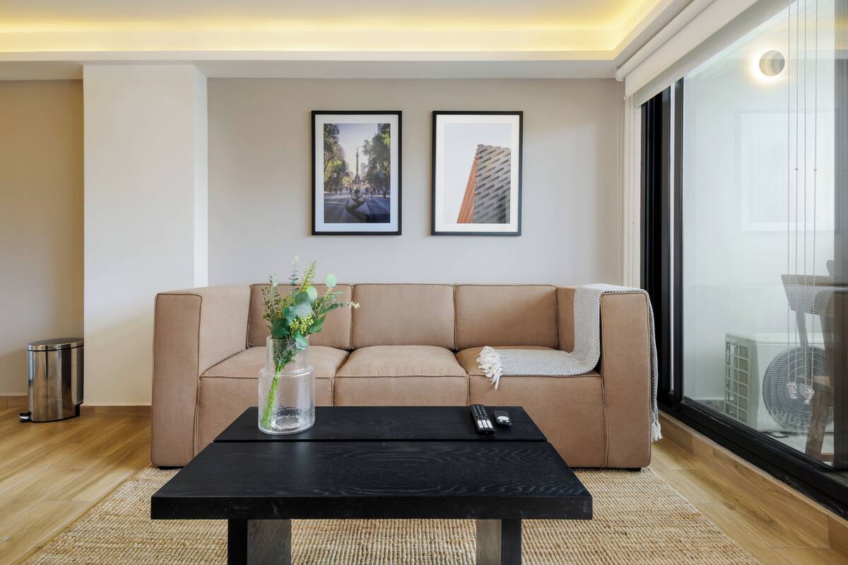 Uma sala de estar moderna com um sofá bege, uma mesa de centro preta e duas imagens emolduradas na parede.