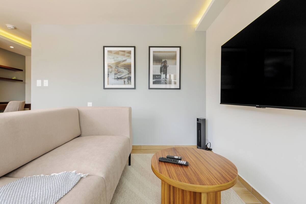 Uma sala de estar moderna com um sofá bege, uma mesa de centro redonda de madeira e uma televisão de tela plana fixada na parede.