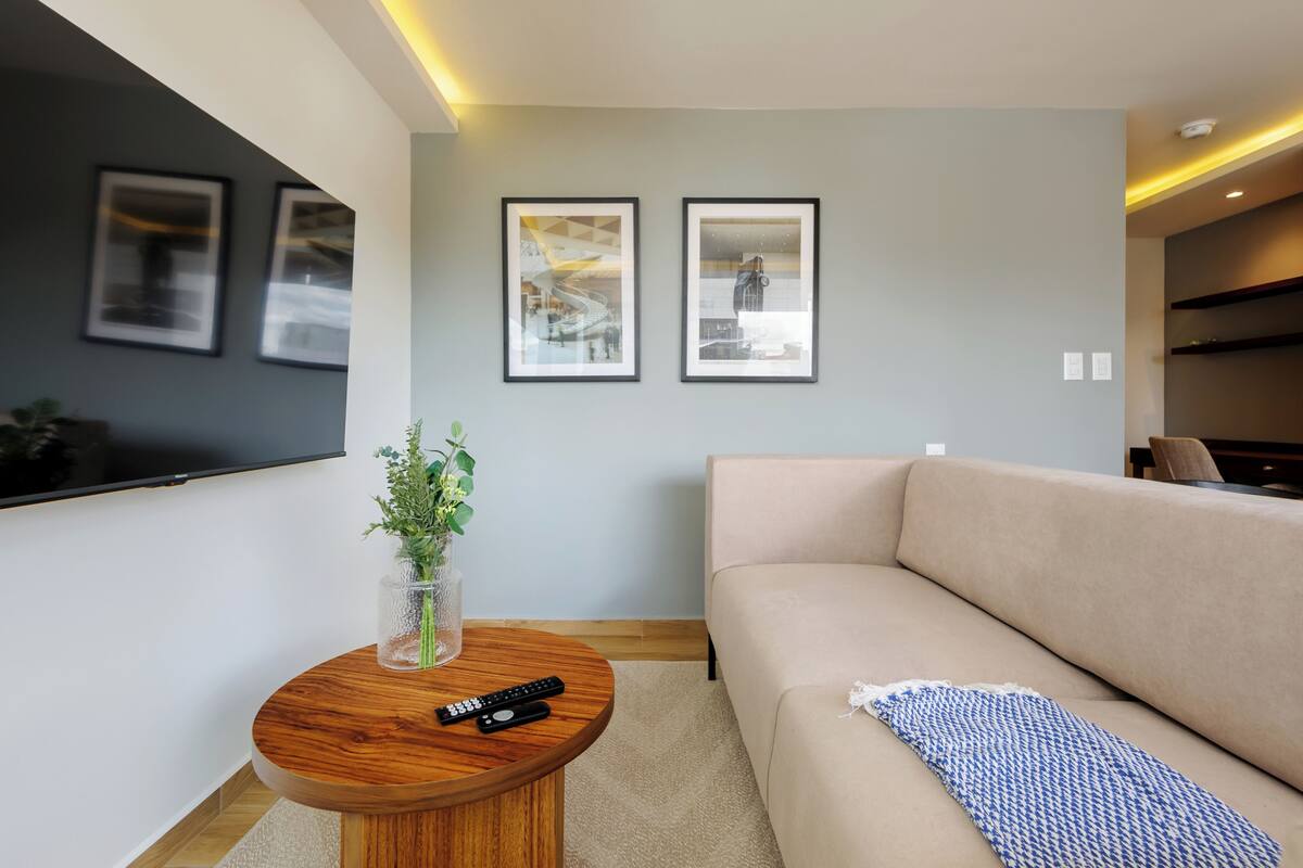 Uma sala de estar moderna com um sofá bege, uma mesa de centro redonda de madeira com um vaso de plantas verdes, e duas fotos emolduradas na parede.