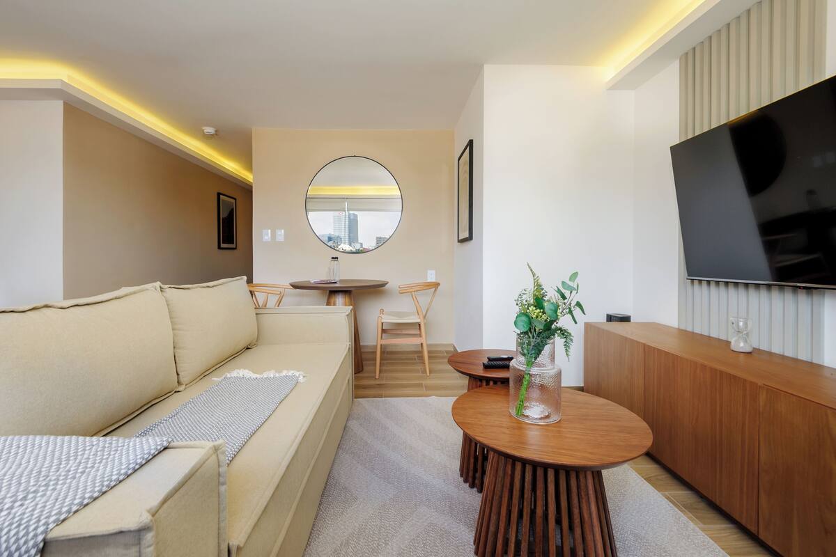 Quarto de hotel moderno com sofá, mesa de jantar redonda, TV e espelho.