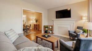 Uma sala de estar com sofá, poltrona, mesa de centro, lareira, TV e um espaço de jantar ao fundo.
