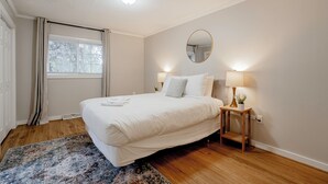 Um quarto bem arrumado com uma cama grande, uma janela com cortinas, uma mesa de cabeceira com abajur e um espelho na parede.