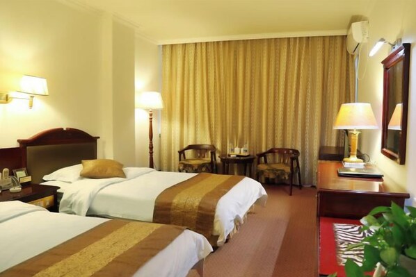 Quarto de hotel com duas camas, uma escrivaninha, uma cadeira, um abajur e uma janela grande com cortinas.