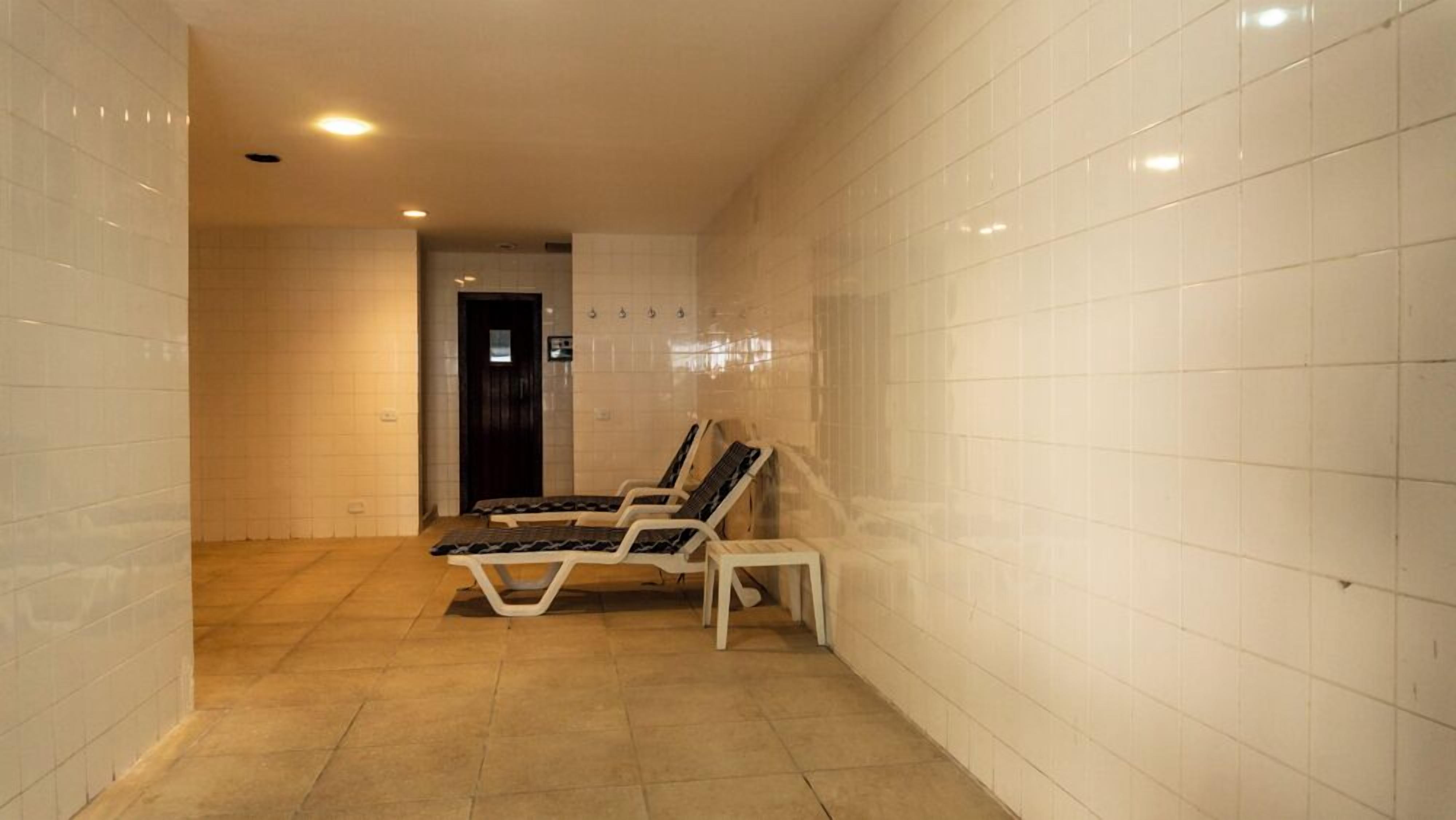 Um corredor com paredes e chão em azulejo branco que leva a um quarto com porta escura. Há duas poltronas de lounge com estrutura branca e almofadas pretas.