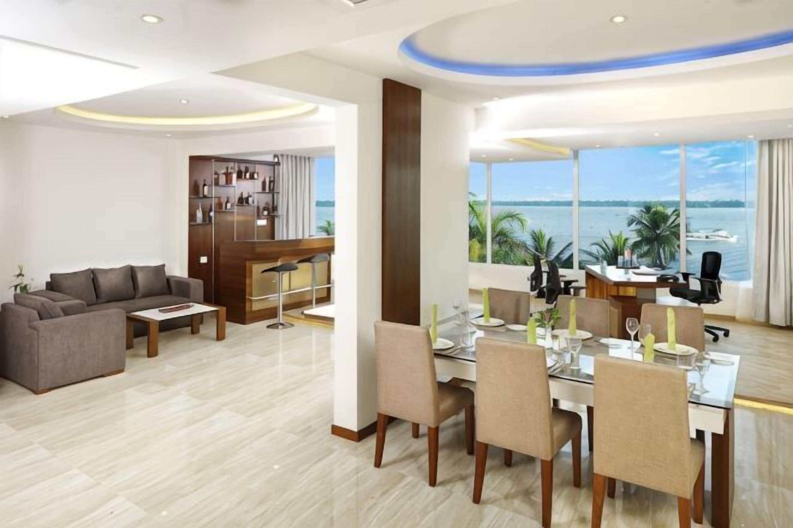 Quarto de hotel moderno com área de jantar, sofá, mesa de centro e vista para o mar.