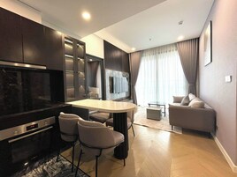 Cozinha moderna com armários escuros, forno embutido e uma televisão grande. Há uma sala de jantar com mesa redonda e cadeiras, e uma sala de estar com sofá e televisão.