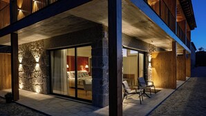 Edifício moderno com fachada de pedra, porta de entrada de vidro que dá para um quarto com cama e mesa de cabeceira, e uma pequena área externa com dois assentos.