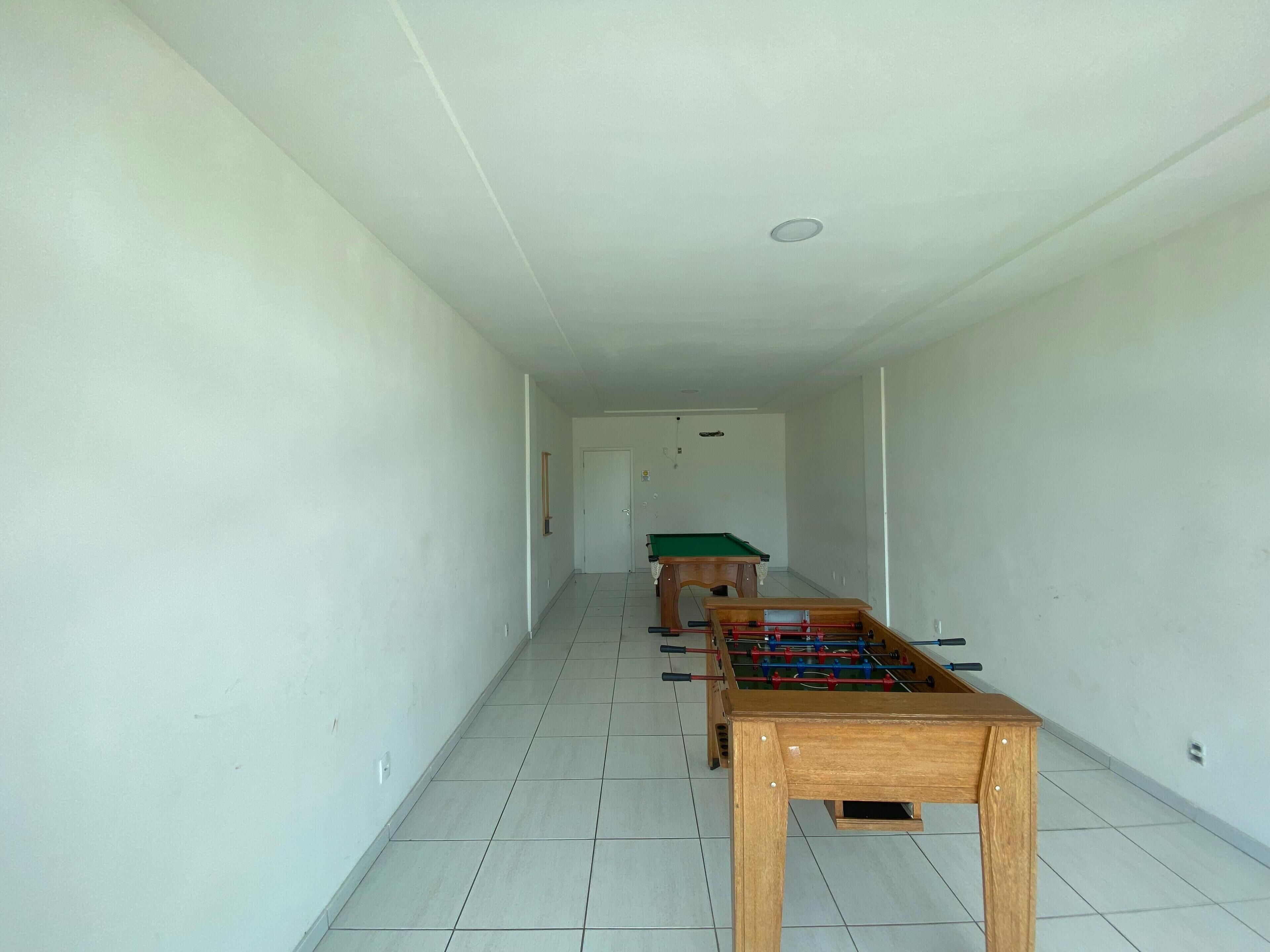 Um corredor com duas mesas de pebolim e uma mesa de bilhar, pisos de azulejos brancos e paredes sem decoração.