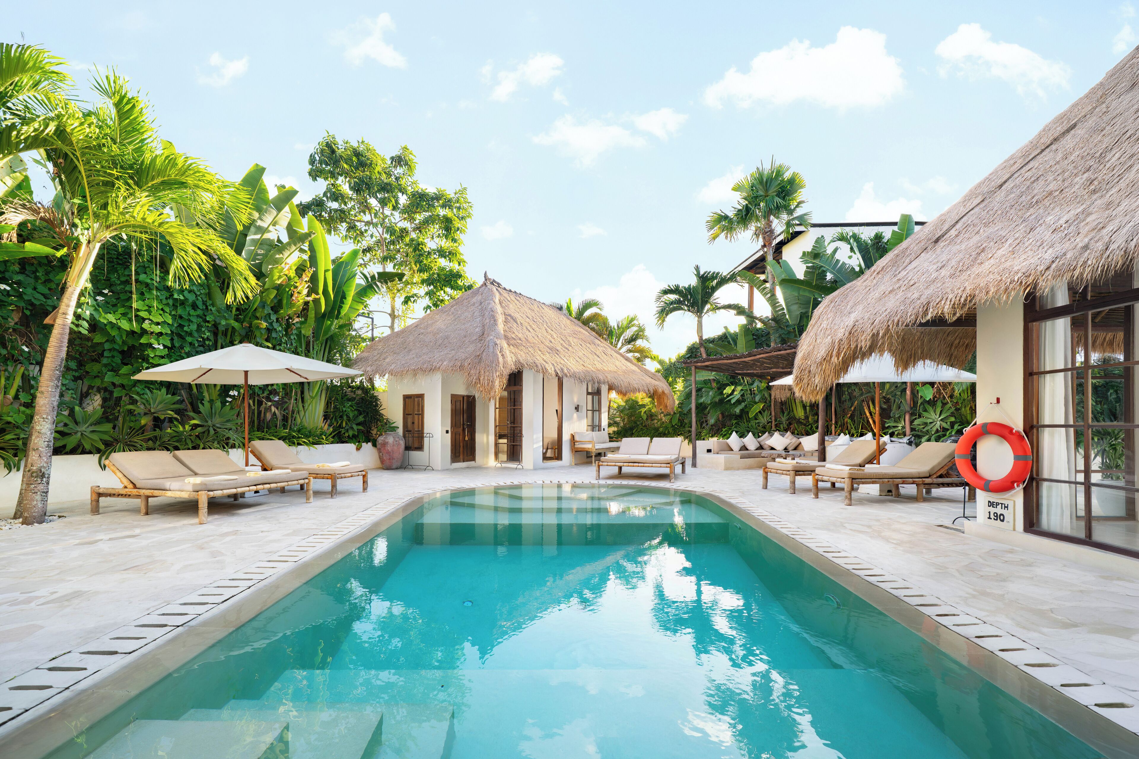 Área de piscina com espreguiçadeiras, uma estrutura com telhado de palha e vegetação tropical.