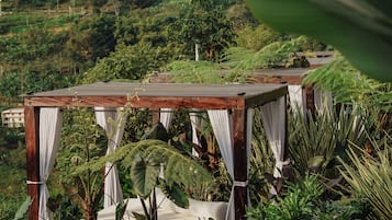 Um gazebo de madeira com cortinas brancas, rodeado por vegetação exuberante e plantas tropicais.