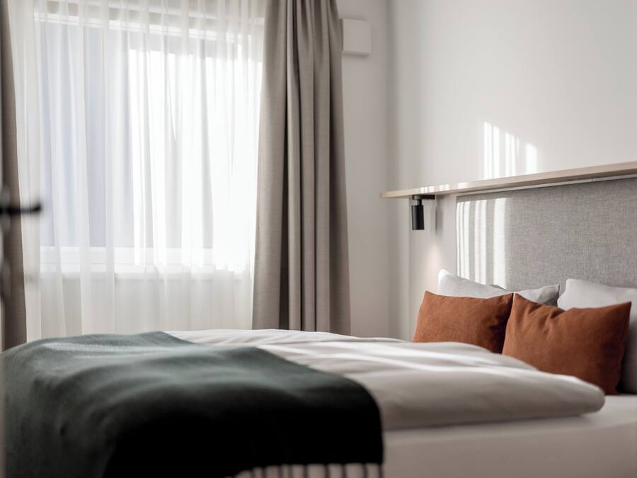 Quarto de hotel moderno com cama bem arrumada, cabeceira e uma grande janela com cortinas leves.