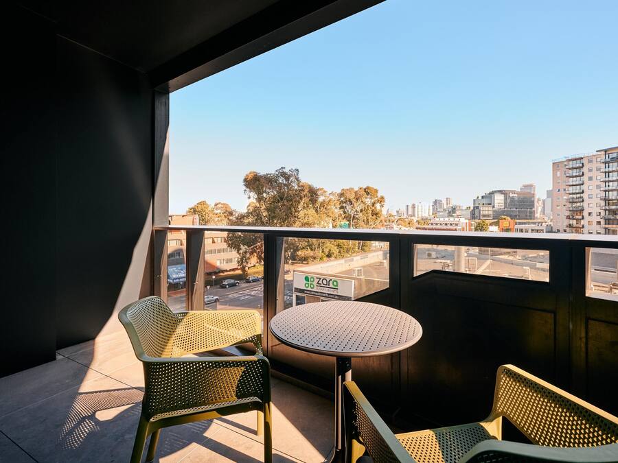 Varanda com duas cadeiras de vime e uma mesa redonda pequena, com vista para um horizonte urbano com edifícios e árvores.