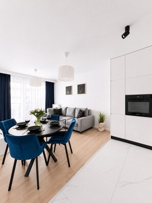 Uma sala de estar moderna com área de jantar, um sofá cinzento, uma cozinha branca com forno e uma planta em vaso.