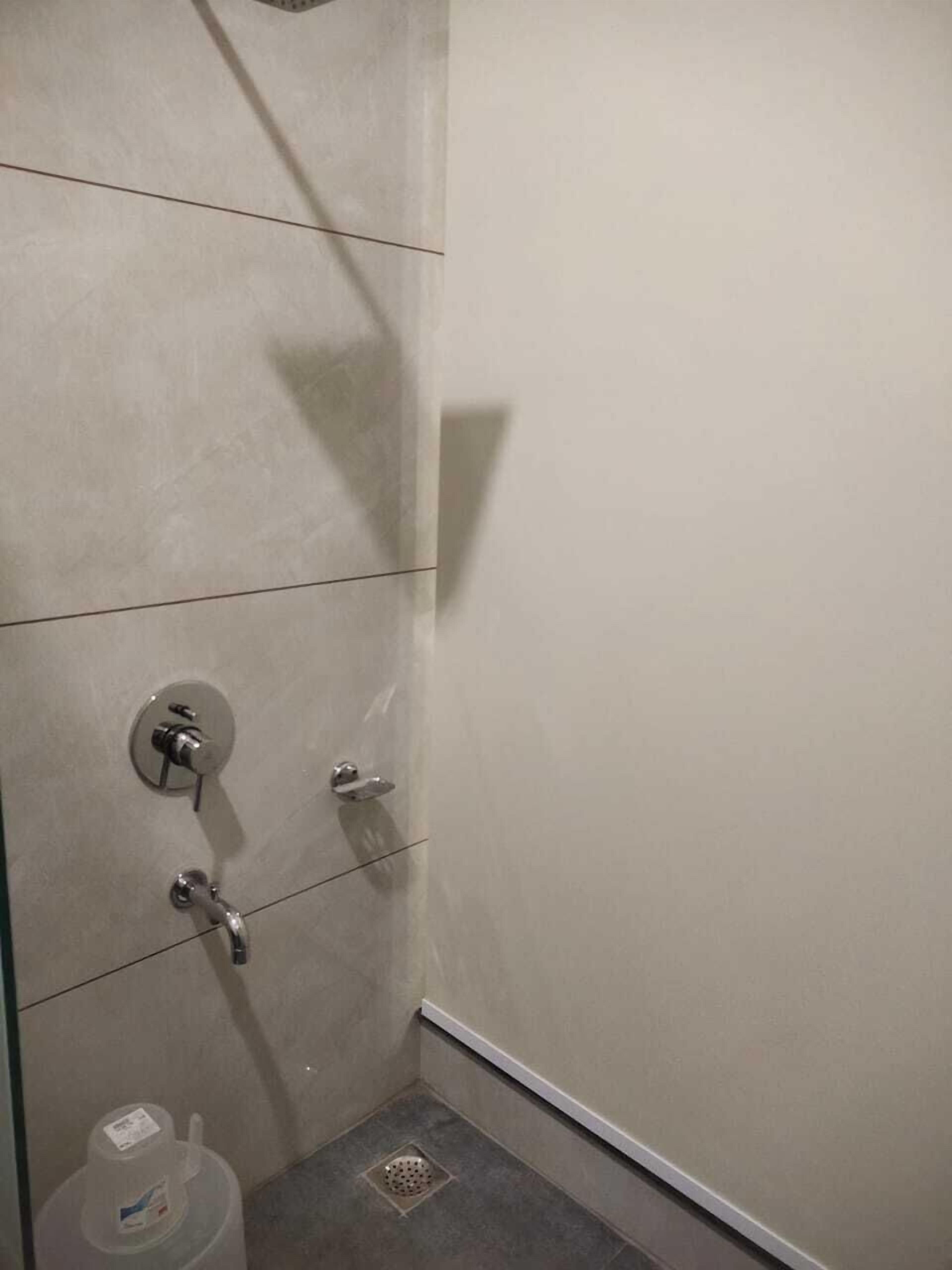 Pequena área de chuveiro revestida com azulejos, com chuveiro manual, torneira embutida na parede e ralo.