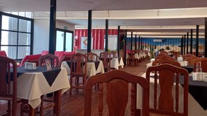 Restaurante com mesas e cadeiras de madeira, janelas amplas e um fundo de cortina vermelha e azul.