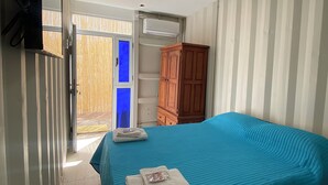 Quarto com colcha azul, guarda-roupa de madeira, ventoinha no teto e janela com vista para uma cerca de bambu.