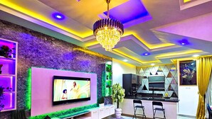 Uma sala de estar moderna com televisão de tela plana, um candelabro e um espaço de jantar com cadeiras, com uma cozinha ao fundo.