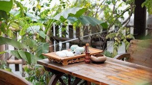 Uma mesa de madeira com uma bandeja de xícaras de chá, um bule e uma tigela de madeira, disposta ao ar livre com vegetação exuberante ao fundo.
