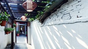 Um beco estreito com uma pérgola de madeira acima, plantas em vasos à esquerda e um muro de tijolos brancos à direita.